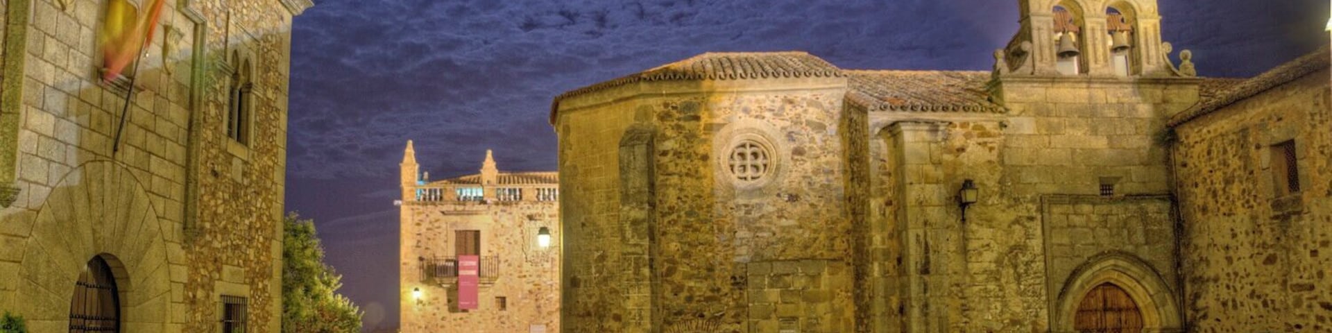 Um edifício histórico de pedra com uma torre de sino, rodeado por ruas de calçada e plantas em vasos.