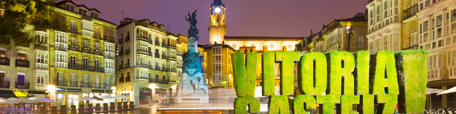 Praça urbana iluminada à noite, com edifícios, uma estátua e o texto 'VITORIA CASTELLO'.