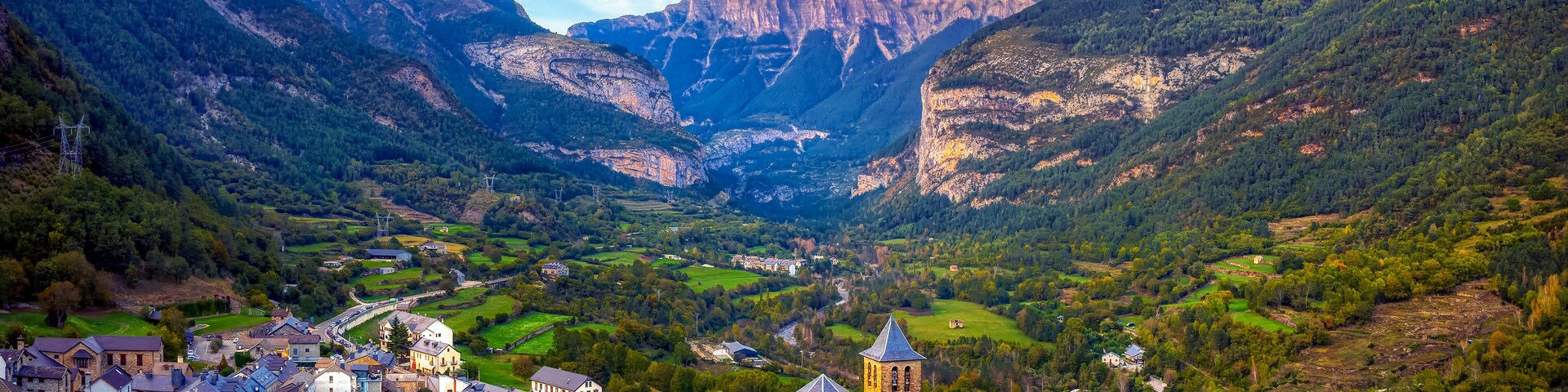 Paisagem montanhosa com uma aldeia aninhada num vale, destacando-se uma torre de igreja e vegetação exuberante.