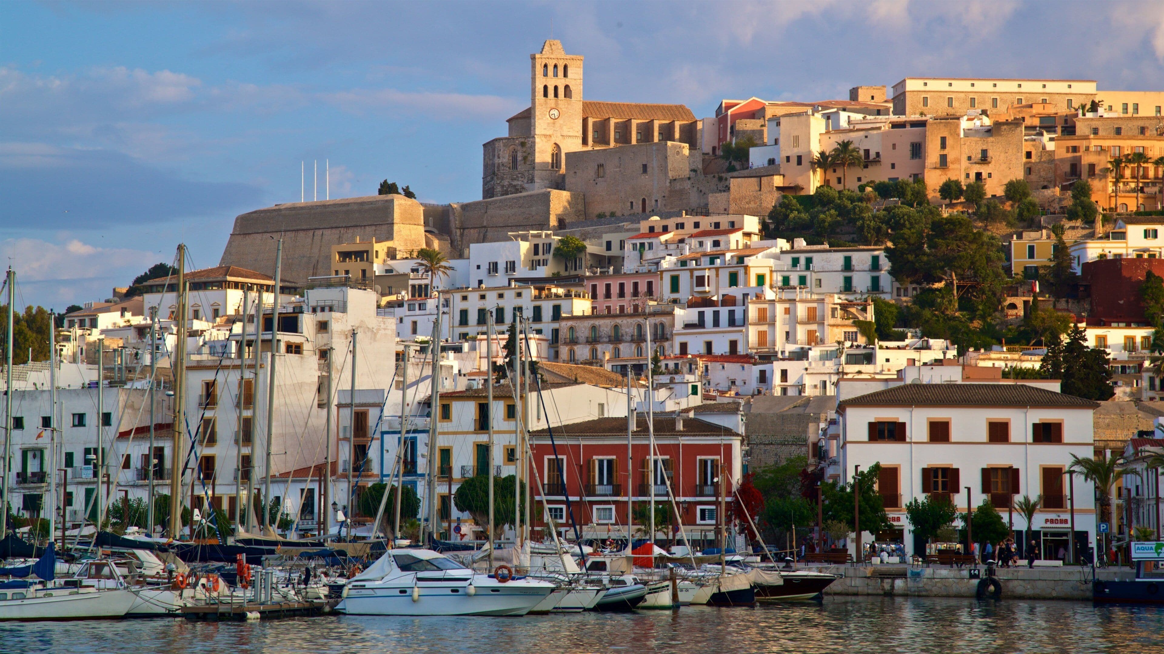 Uma cidade costeira com edifícios históricos, uma torre imponente e uma marina com barcos.