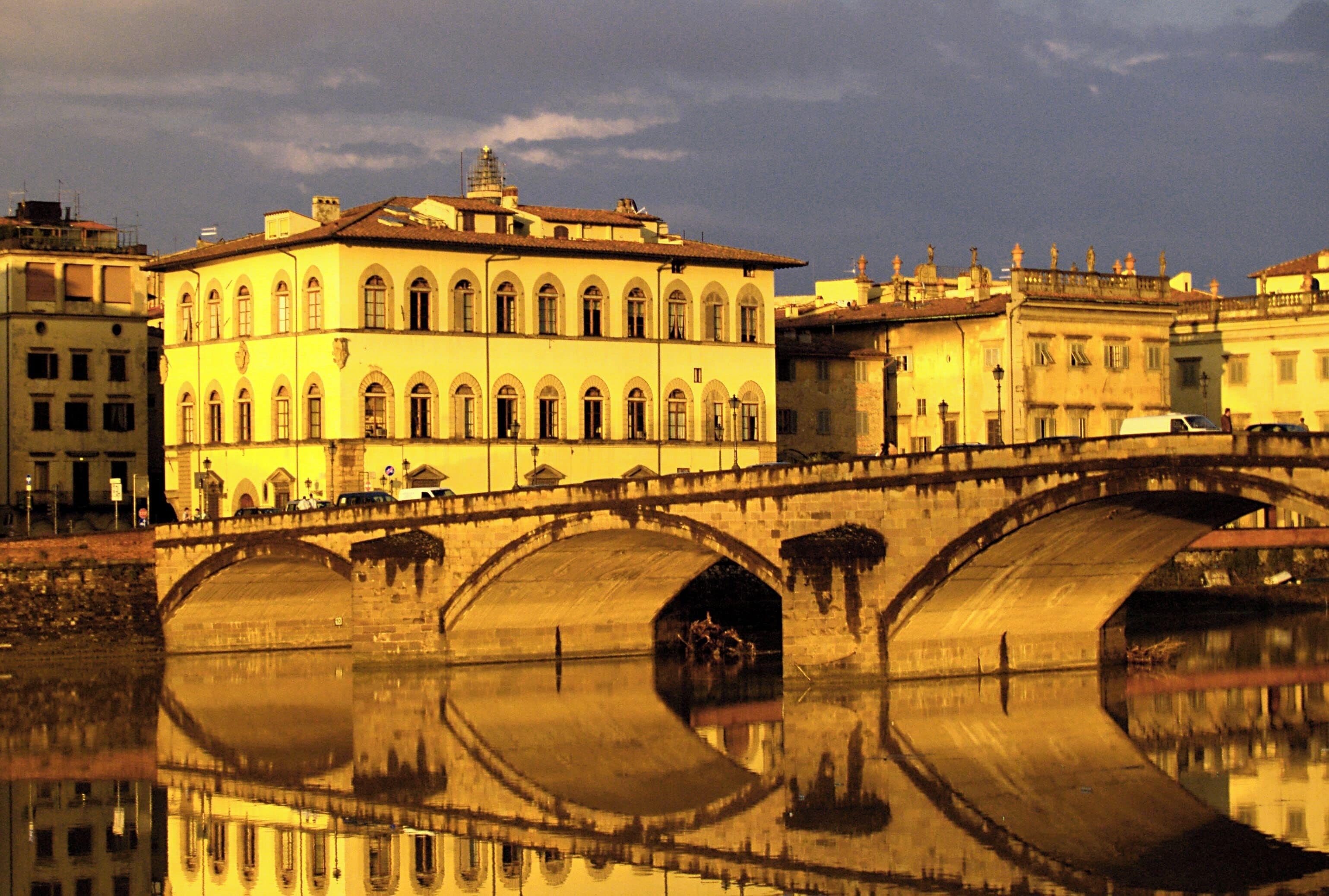 Uma ponte com arcos sobre a água, refletindo edifícios do outro lado.