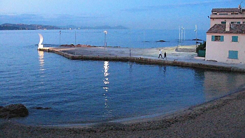 Uma orla calma com um cais que se estende sobre a água, um pequeno edifício com telhado de azulejos e algumas pessoas caminhando pela margem.