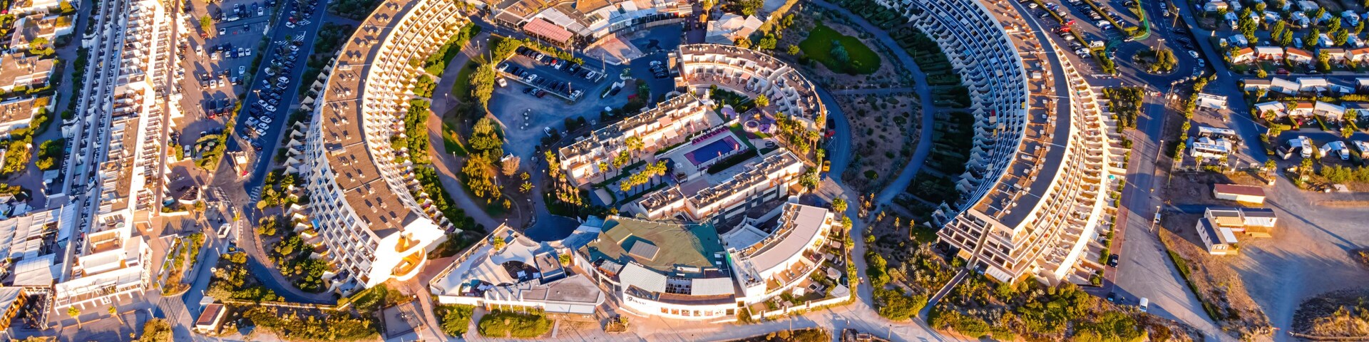 Vista aérea de uma cidade costeira com um porto circular, uma praia com espreguiçadeiras e uma área residencial densa.