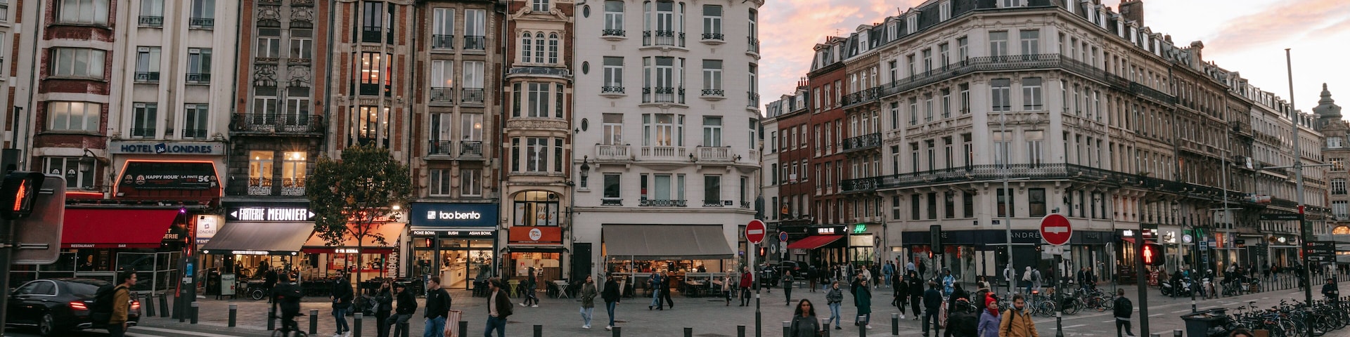 Uma rua urbana movimentada com arquitetura europeia histórica, incluindo um hotel e diversas lojas.