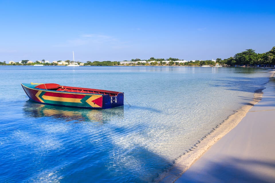 Um barco colorido em águas calmas próximo a uma praia arenosa, com uma paisagem urbana ao longe.