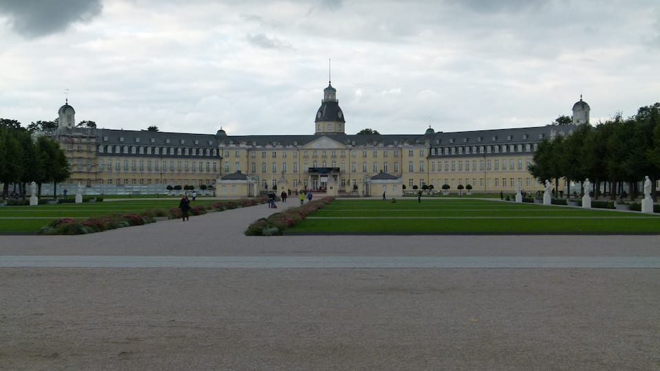 Um edifício histórico imponente, com arquitetura simétrica, uma cúpula central e um jardim formal com estátuas.