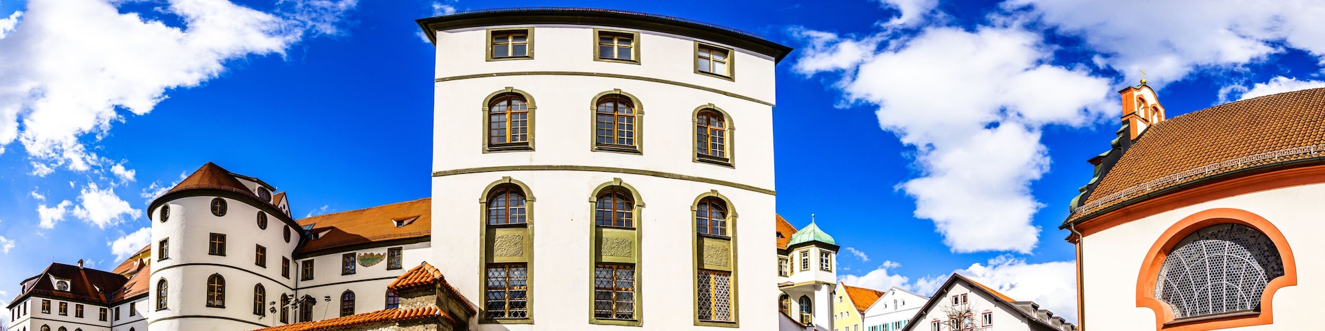 Um edifício histórico com fachada branca e janelas em arco, rodeado por outros edifícios de arquitetura europeia tradicional.