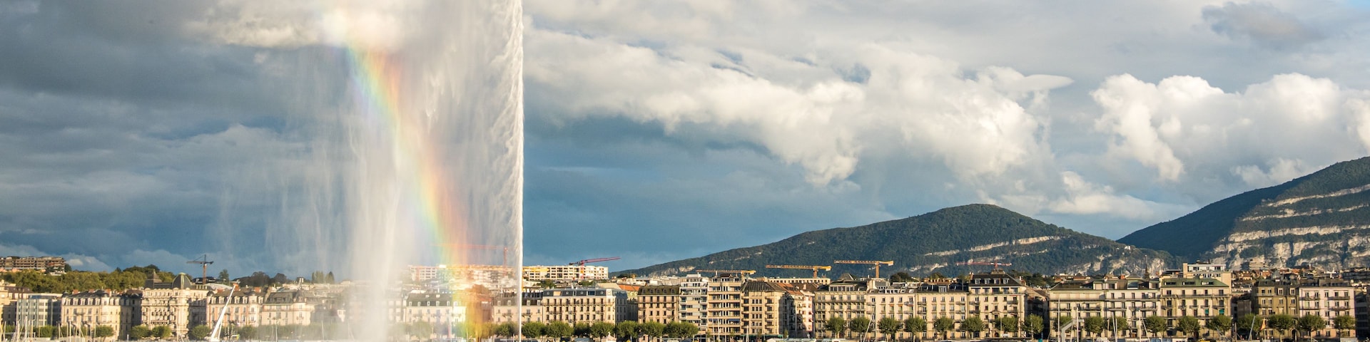 Paisagem urbana com uma fonte, um arco-íris e um barco na água.