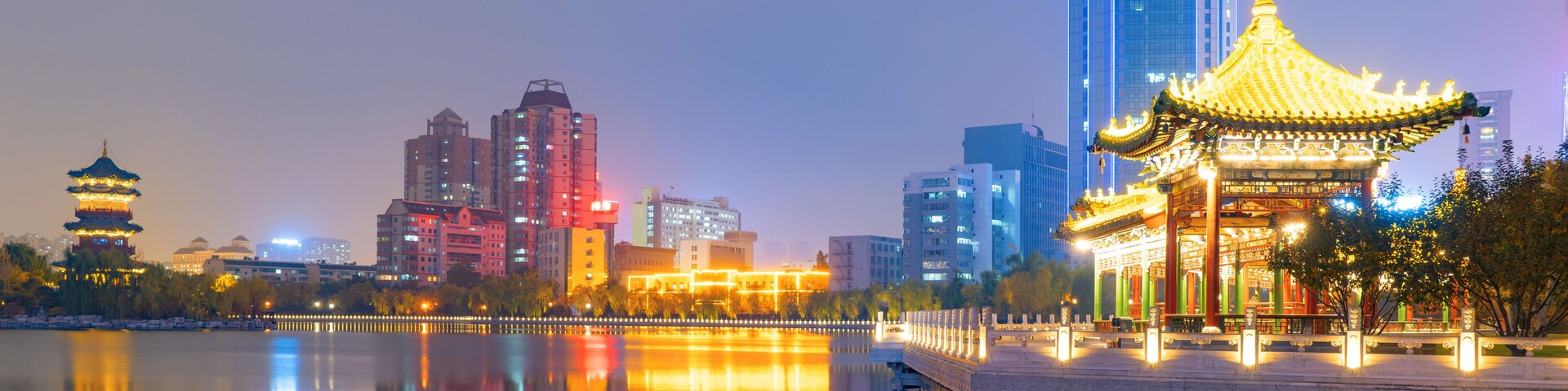 Paisagem urbana ao entardecer com edifícios iluminados, um pagode tradicional e um rio calmo refletindo as luzes.