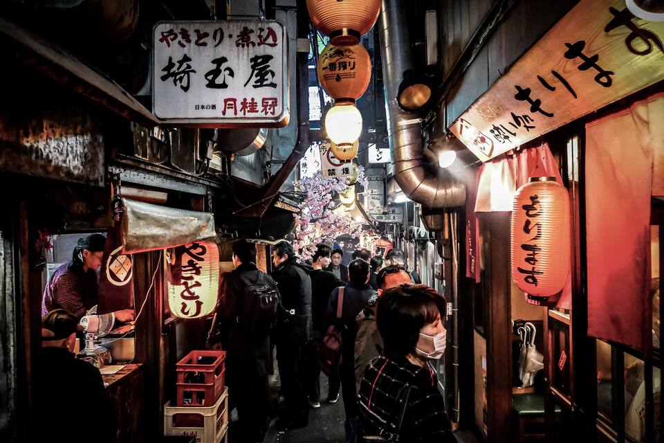 Uma viela estreita com sinalização japonesa tradicional, lanternas e pessoas caminhando.