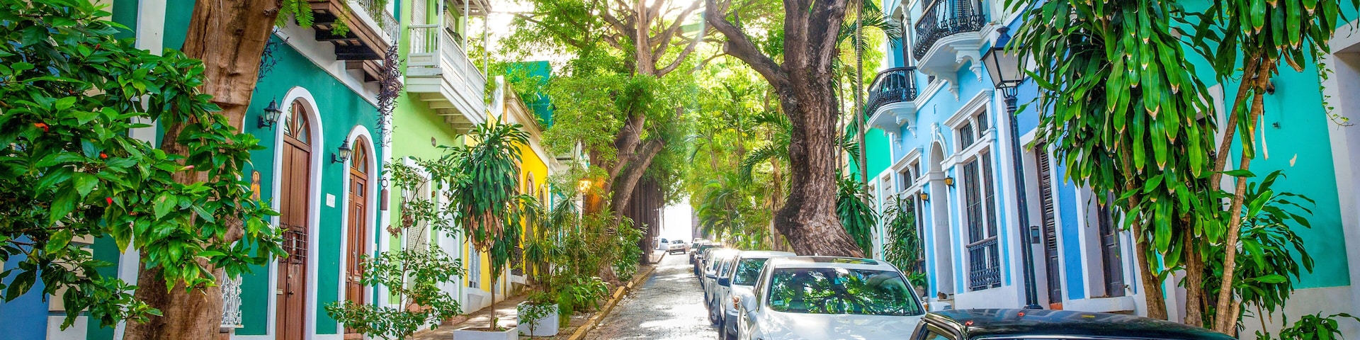 Uma rua de paralelepípedos ladeada por edifícios coloridos, carros estacionados e vegetação exuberante.