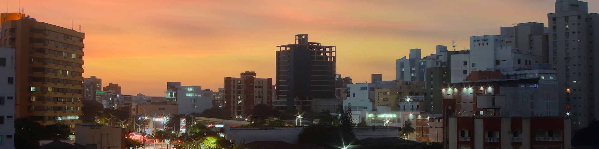 Paisagem urbana ao entardecer com céu avermelhado, edifícios e rua movimentada com rastros de luz de veículos.