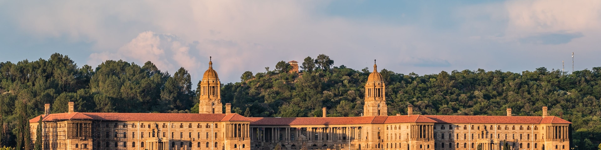 Um edifício histórico imponente com duas torres marcantes, rodeado por vegetação exuberante e um cenário de colinas.