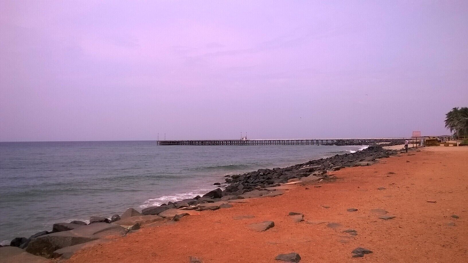 Uma praia com areia avermelhada, um trecho de costa rochosa e um píer que se estende sobre a água.