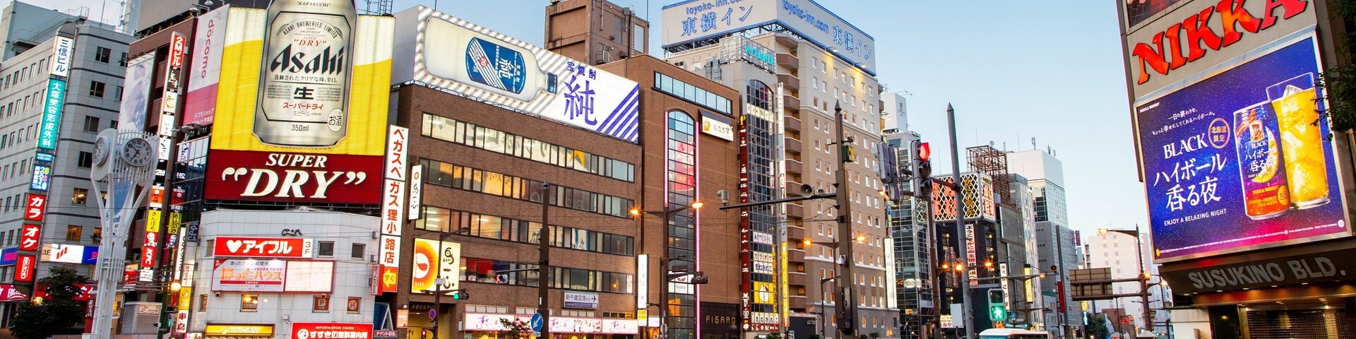 Uma rua urbana movimentada com diversos edifícios comerciais, incluindo um destaque para a placa 'Nikko' e um anúncio de cerveja 'Super Dry'.