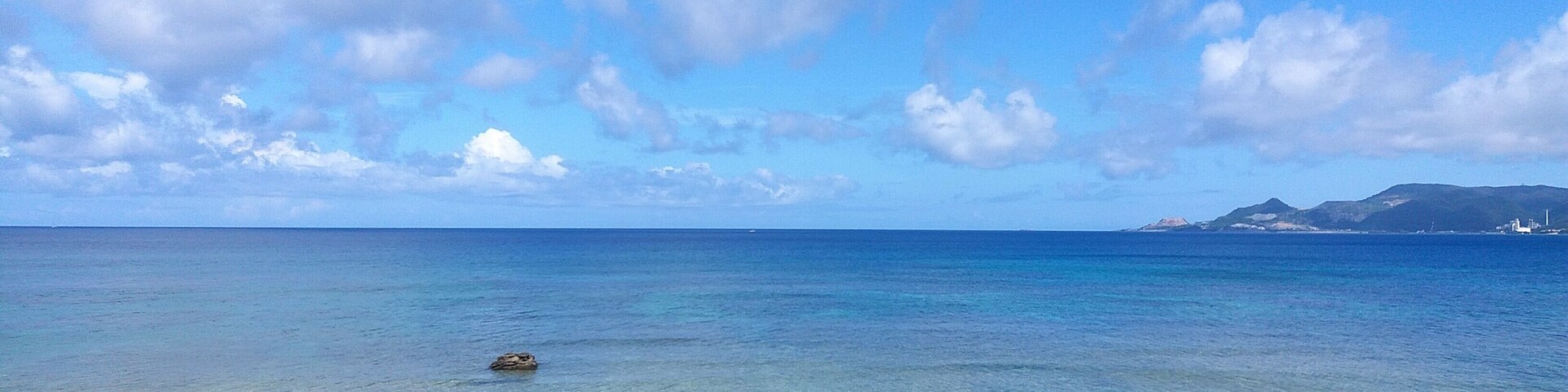 Uma praia tropical tranquila com águas calmas, uma pequena ilha e montanhas ao longe.