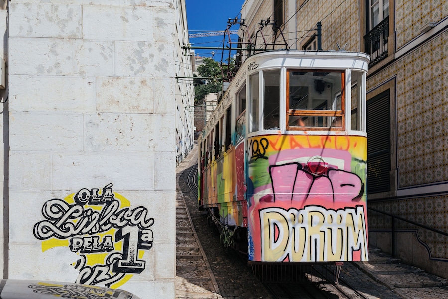 Um elétrico colorido com grafite na lateral, percorrendo uma rua estreita com edifícios de ambos os lados.