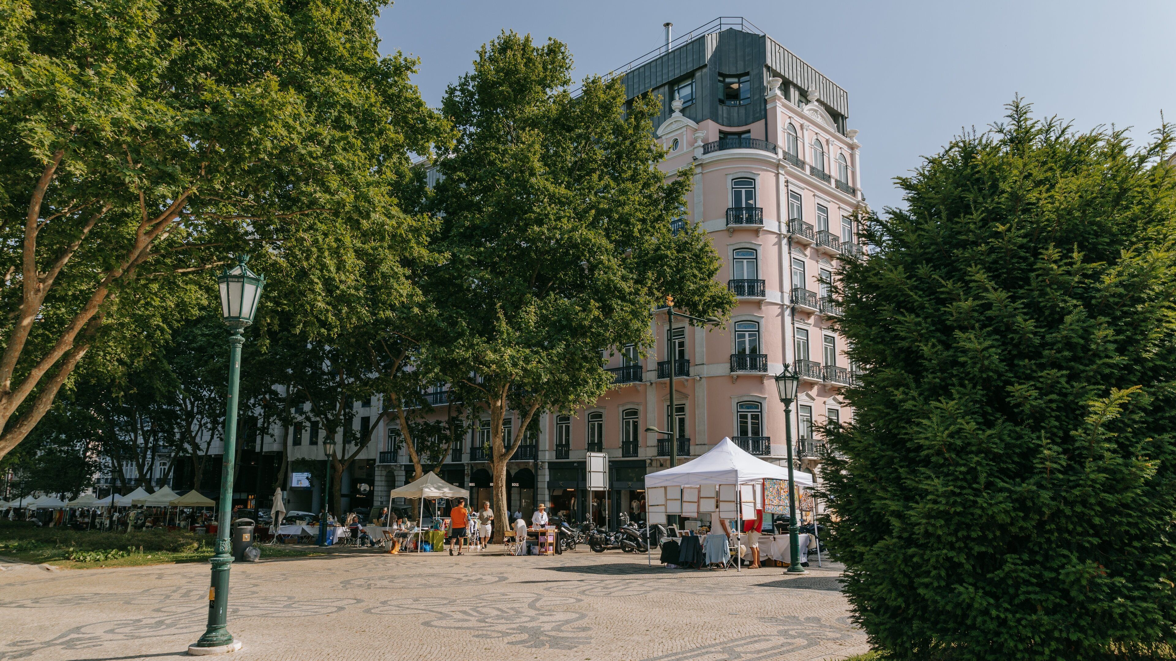 Uma praça de pedras com árvores, uma tenda branca e um edifício rosa com varandas.