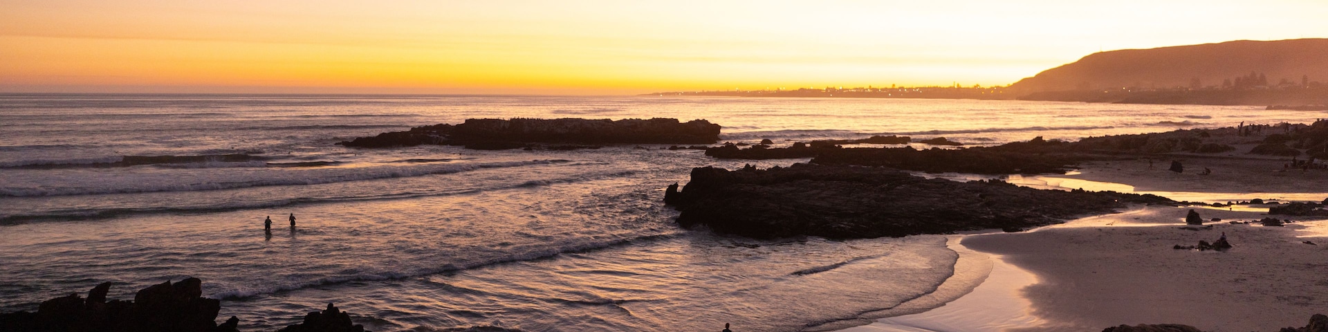 Praia ao pôr do sol com ondas, rochas e o horizonte de uma cidade distante.