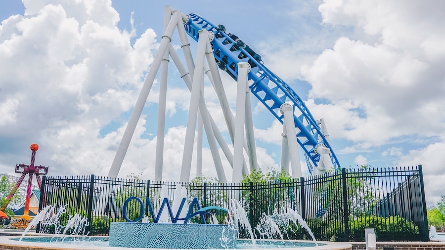 Uma montanha-russa com suportes brancos e uma pista azul, uma fonte de água e uma cerca com as letras "OWA" em primeiro plano.