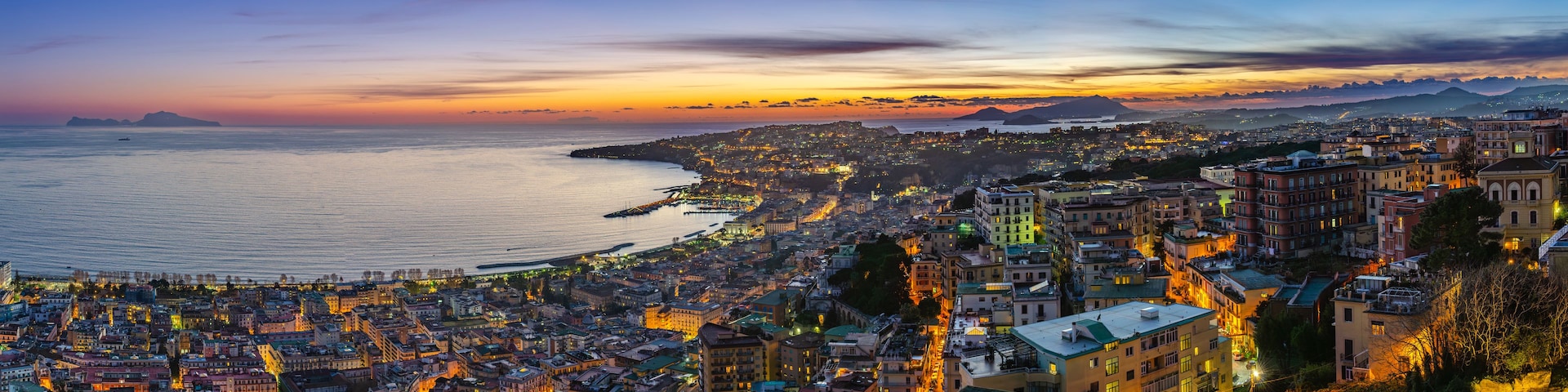 Paisagem costeira ao entardecer com vista para o mar, edifícios iluminados e montanhas ao longe.