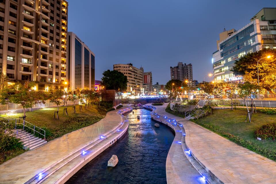 Cena urbana noturna com um espelho de água, caminhos iluminados e edifícios modernos.