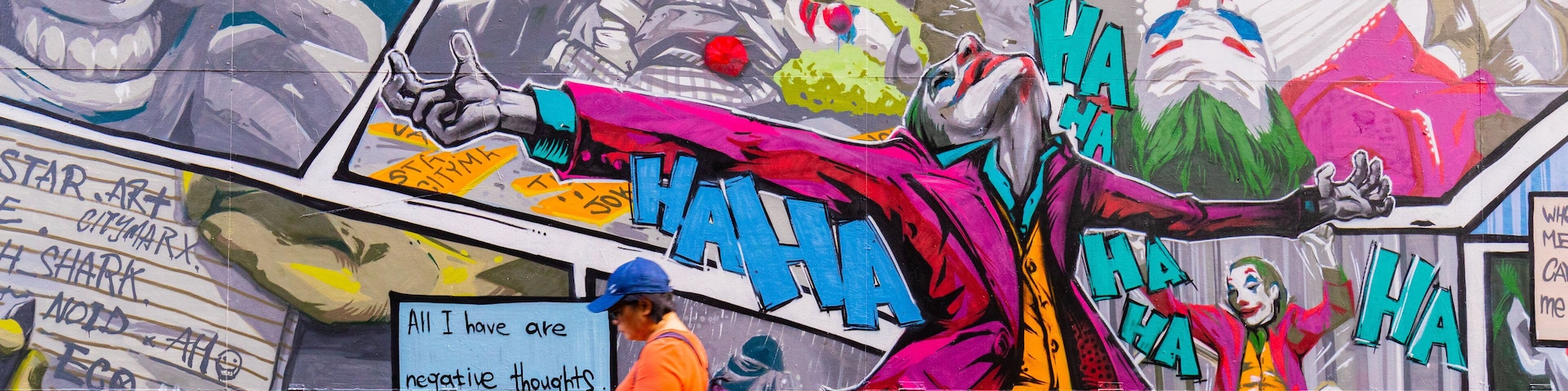 Um mural colorido na rua com arte de graffiti, incluindo a figura de alguém com capa vermelha e a frase 'HA HA' repetida.