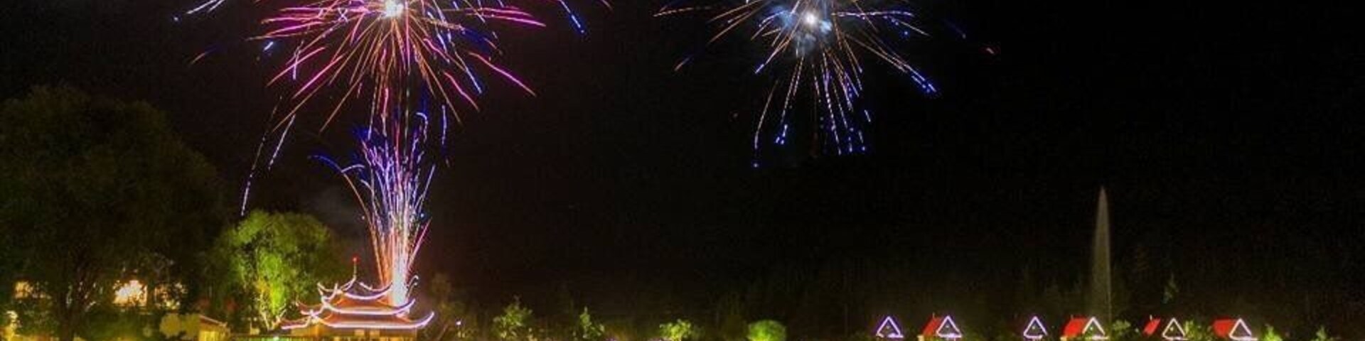 Fogos de artifício sobre um lago tranquilo com edifícios iluminados e um pagode tradicional.
