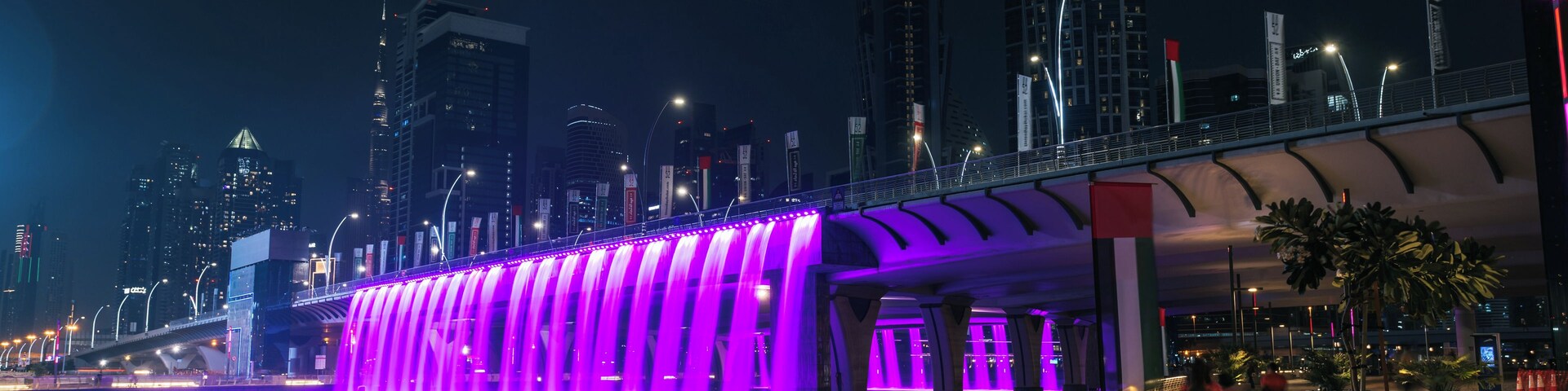 Paisagem urbana noturna com uma ponte e um espelho de água iluminado em roxo.