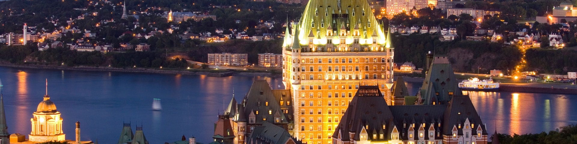 Paisagem urbana ao entardecer, com edifícios iluminados, um rio e uma estrutura imponente semelhante a um castelo com telhado verde.