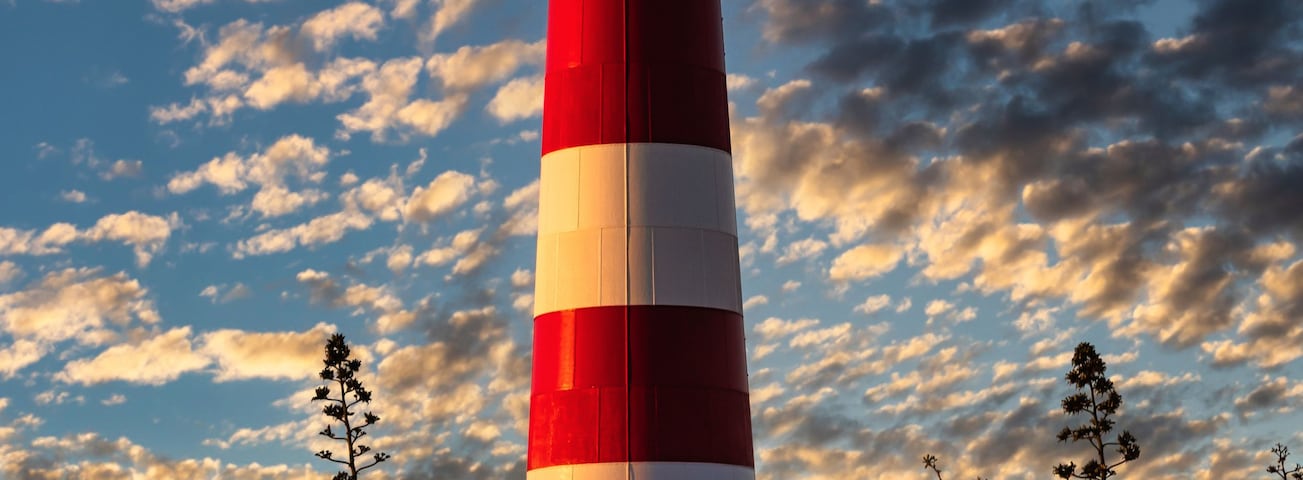 Um farol alto, vermelho e branco, com uma girandola no topo, rodeado por vegetação densa e sob um céu parcialmente nublado.