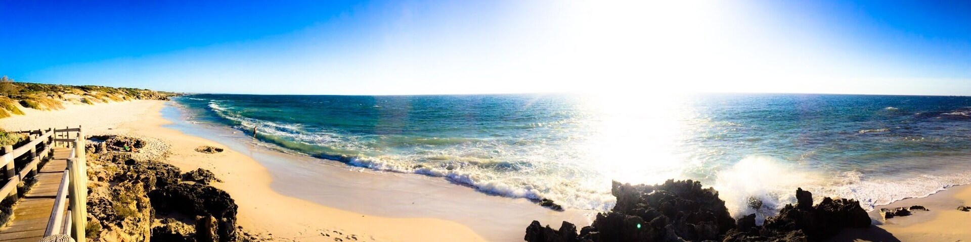 Uma praia com um calçadão de madeira, ondas quebrando na costa e afloramentos rochosos.
