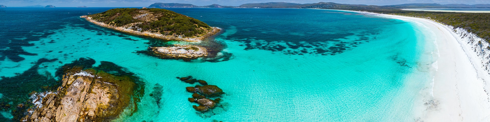 Mar turquesa com praias de areia branca, afloramentos rochosos e montanhas ao longe.