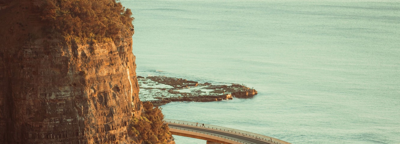 Uma estrada costeira sinuosa serpenteia por um penhasco, com uma ponte sobre a água.