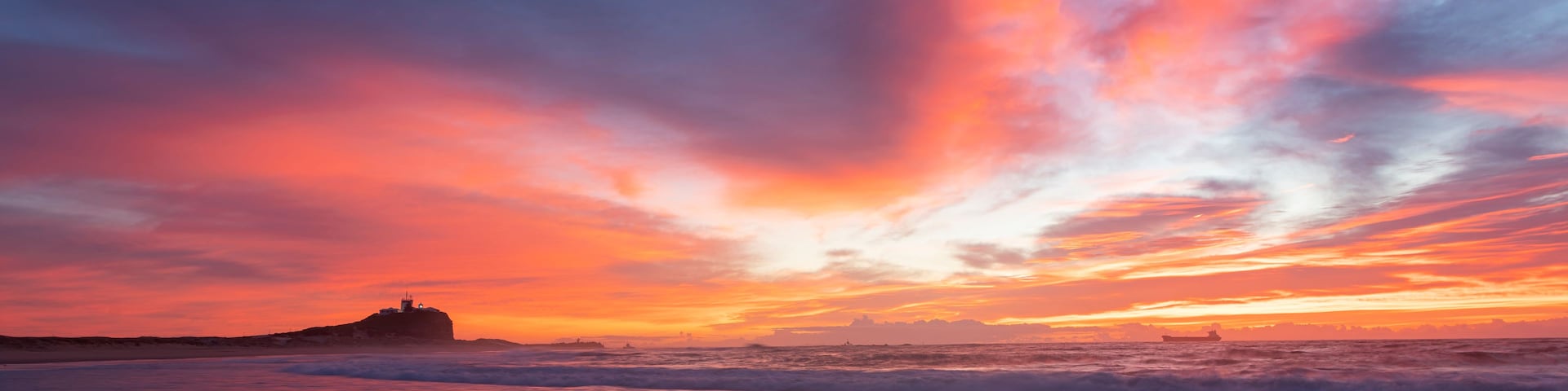 Pôr do sol sobre uma praia com ondas, um navio ao longe e um afloramento rochoso.