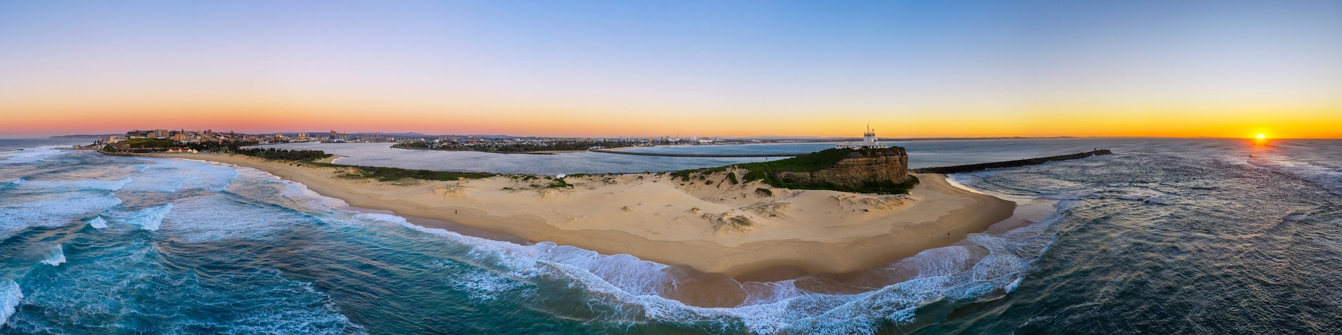 Paisagem costeira com praia arenosa, ondas, um farol e o horizonte da cidade ao longe ao pôr do sol.