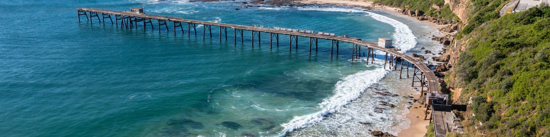 Paisagem costeira com um longo cais que se estende para o mar, uma praia de areia, falésias rochosas e um céu claro.