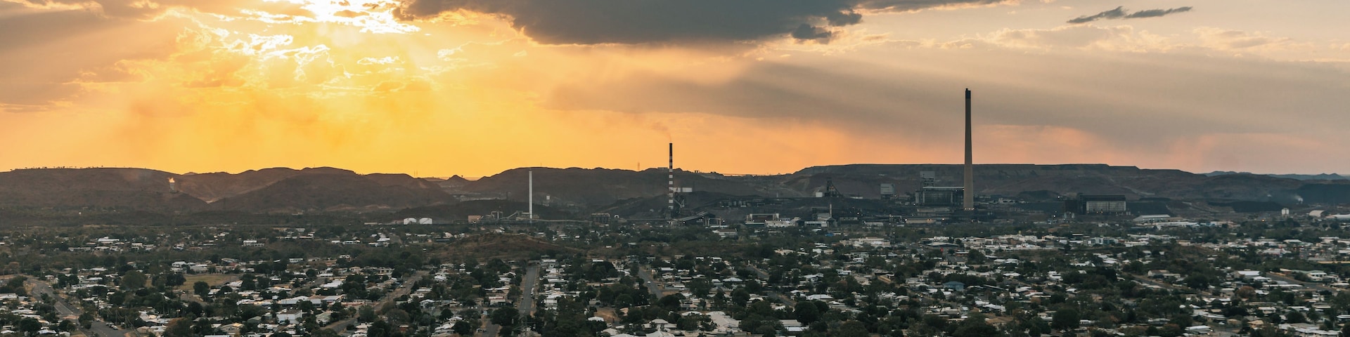 Paisagem urbana com área industrial em destaque, incluindo chaminés altas e uma grande fábrica, sob um céu de pôr do sol dramático.