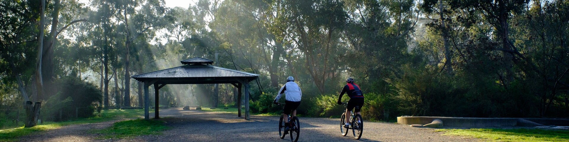 Dois ciclistas em um caminho de cascalho, com um coreto e árvores em um parque.