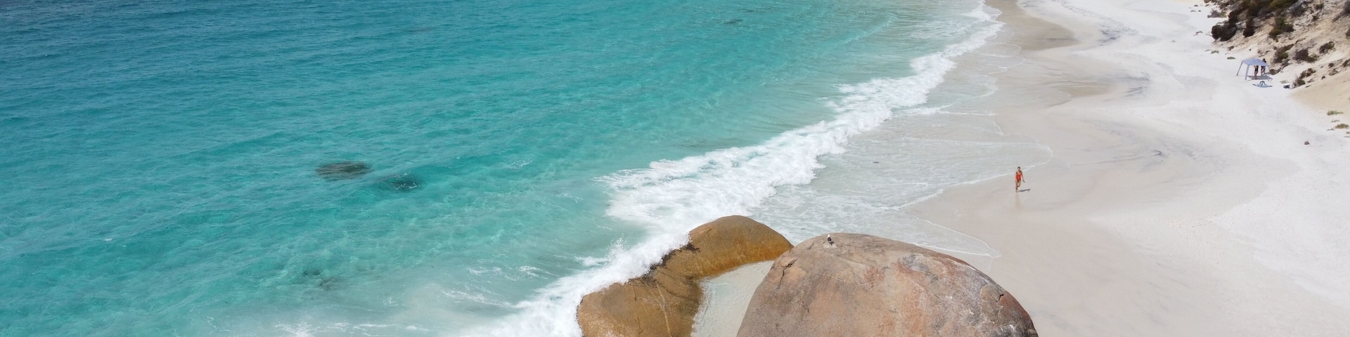 Uma praia com águas turquesa cristalinas, pedras grandes e uma pessoa caminhando na areia.