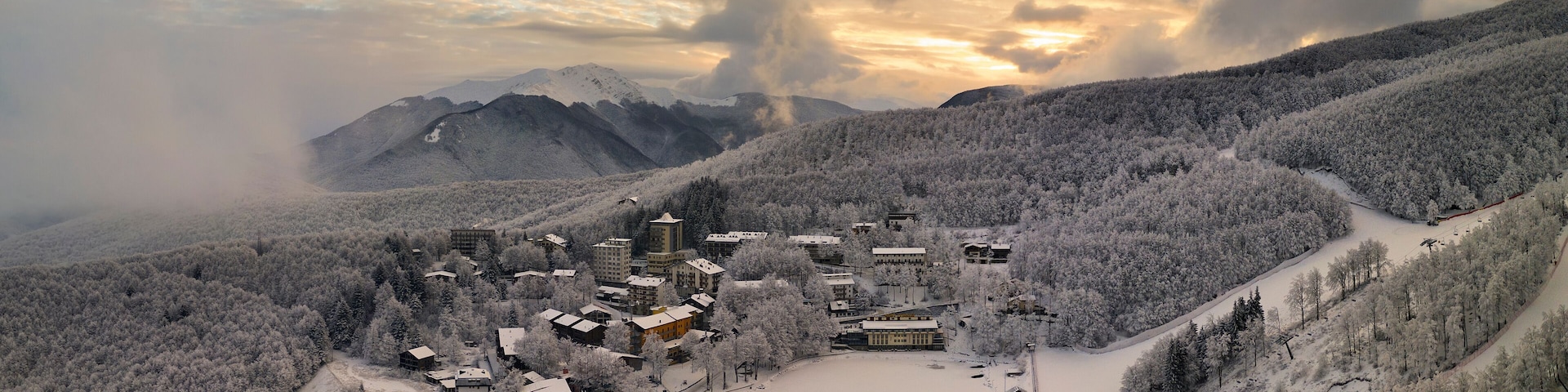 Vila coberta de neve aninhada em uma região montanhosa, com edifícios residenciais e uma grande construção central.