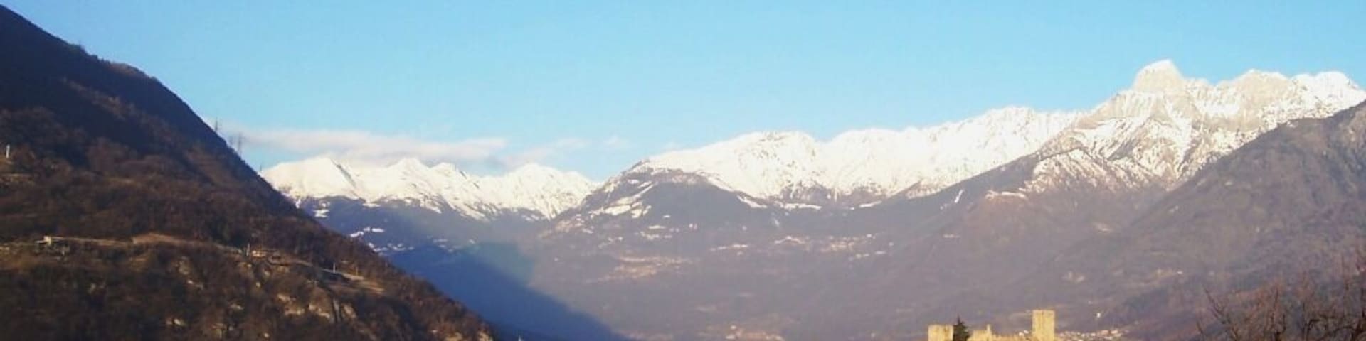 Paisagem montanhosa com um castelo no topo de uma colina, picos nevados ao fundo e céu claro.