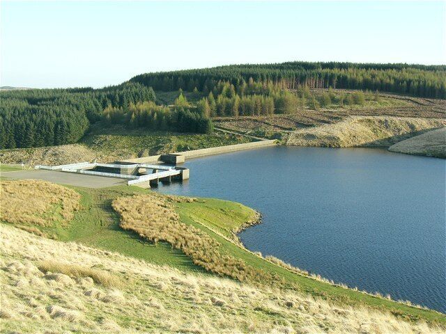 Uma barragem com um reservatório, cercada por colinas arborizadas e um céu claro.