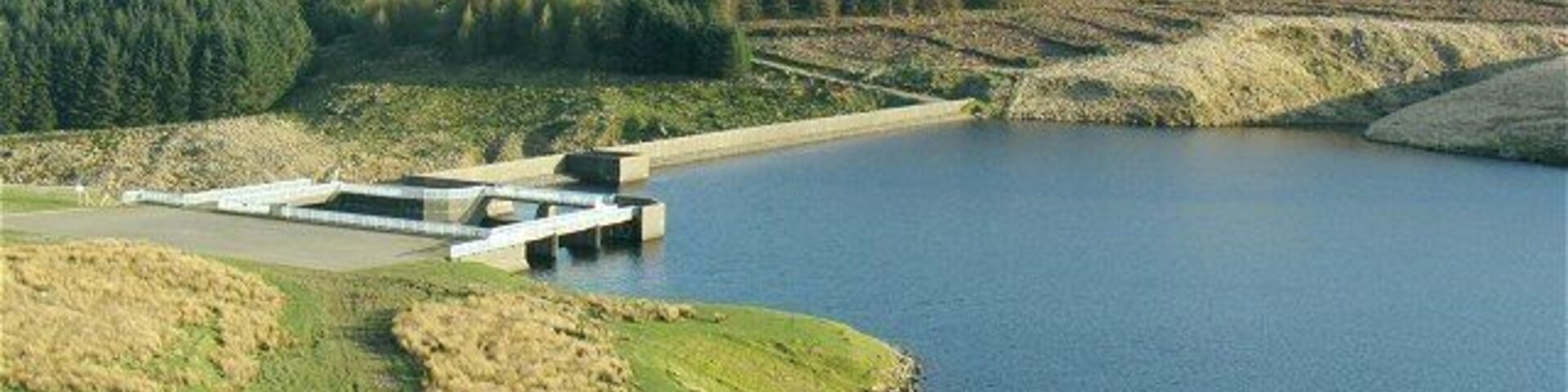 Uma barragem com um reservatório, cercada por colinas arborizadas e um céu claro.