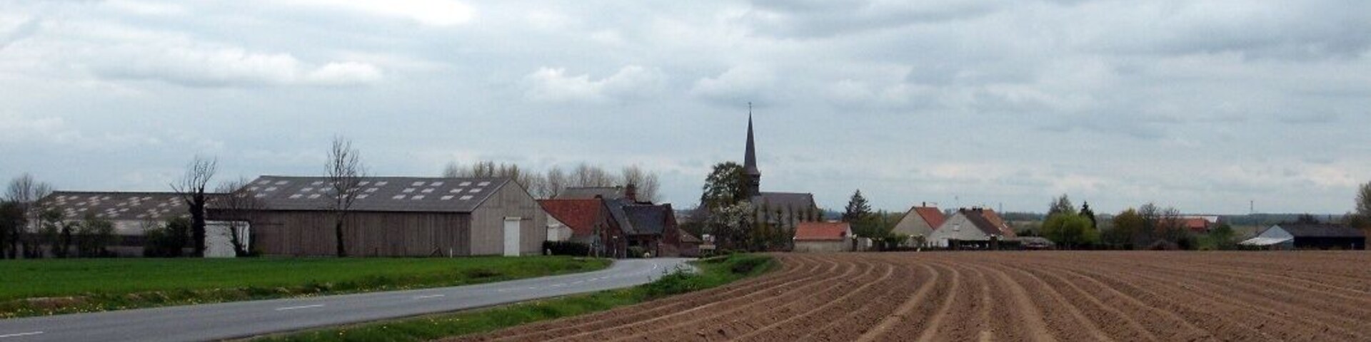 Paisagem rural com estrada serpenteando por campos, uma igreja com torre alta e algumas construções rurais.
