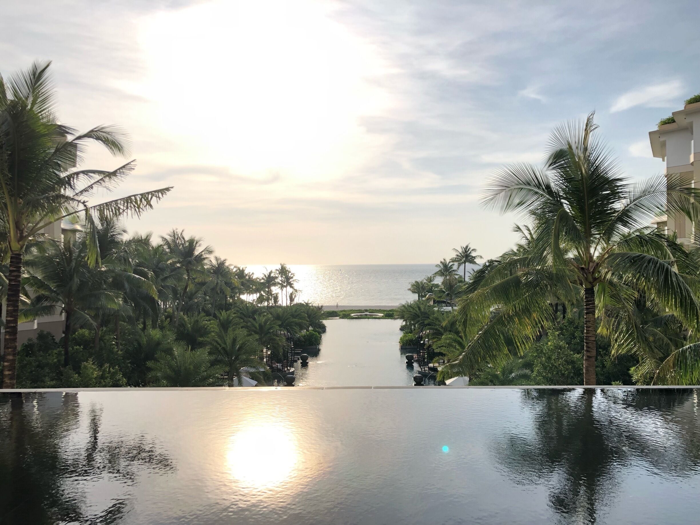 Uma piscina infinita tranquila, com palmeiras e vista para o oceano ao pôr do sol.