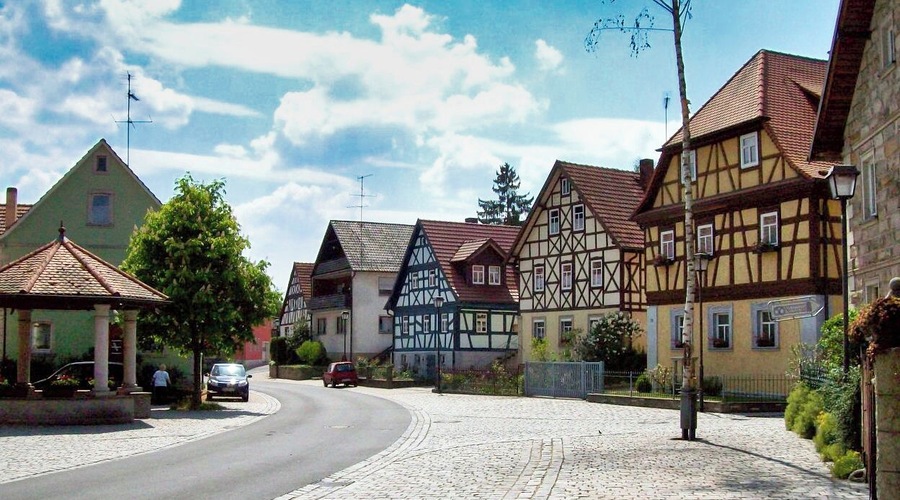 Uma rua de paralelepípedos ladeada por casas tradicionais de enxaimel, um carro estacionado na lateral e um céu azul límpido.