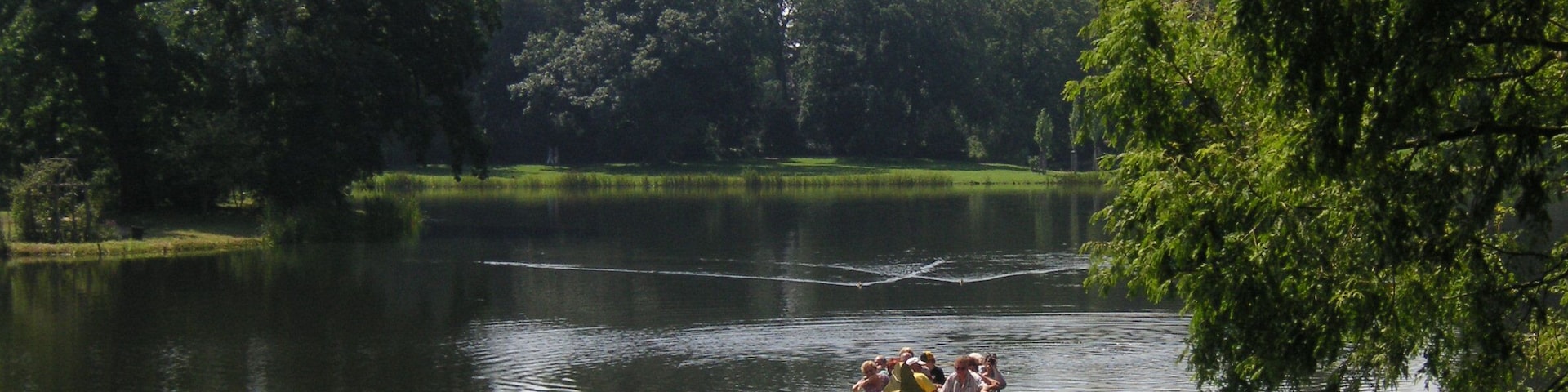 Um lago tranquilo com um barco levando pessoas, cercado por vegetação exuberante e uma torre de igreja ao longe.