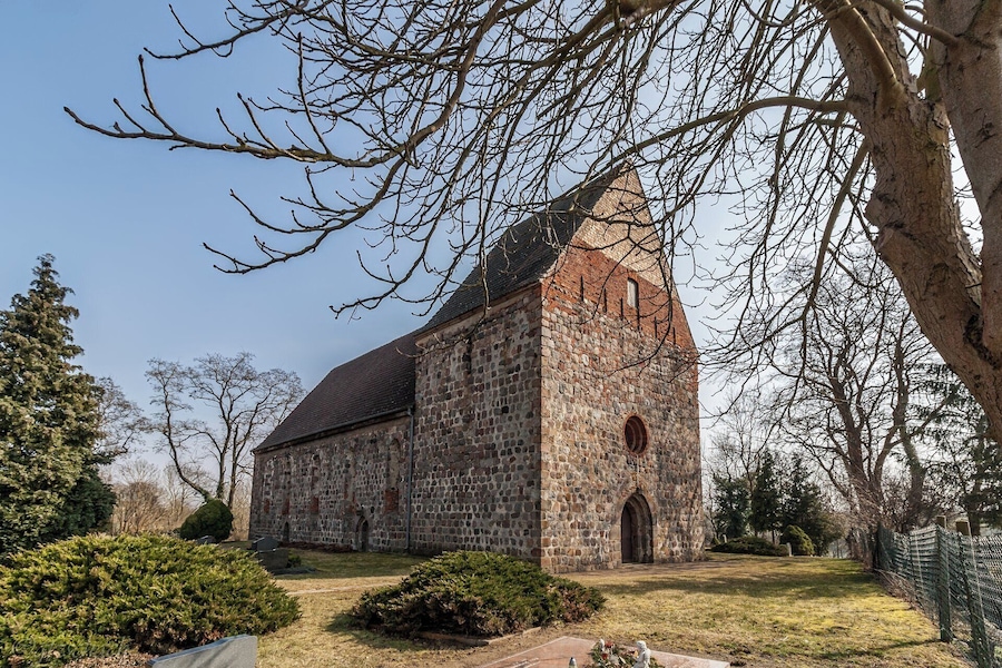 Uma igreja de pedra com uma alta torre sineira, rodeada por árvores e uma área gramada.