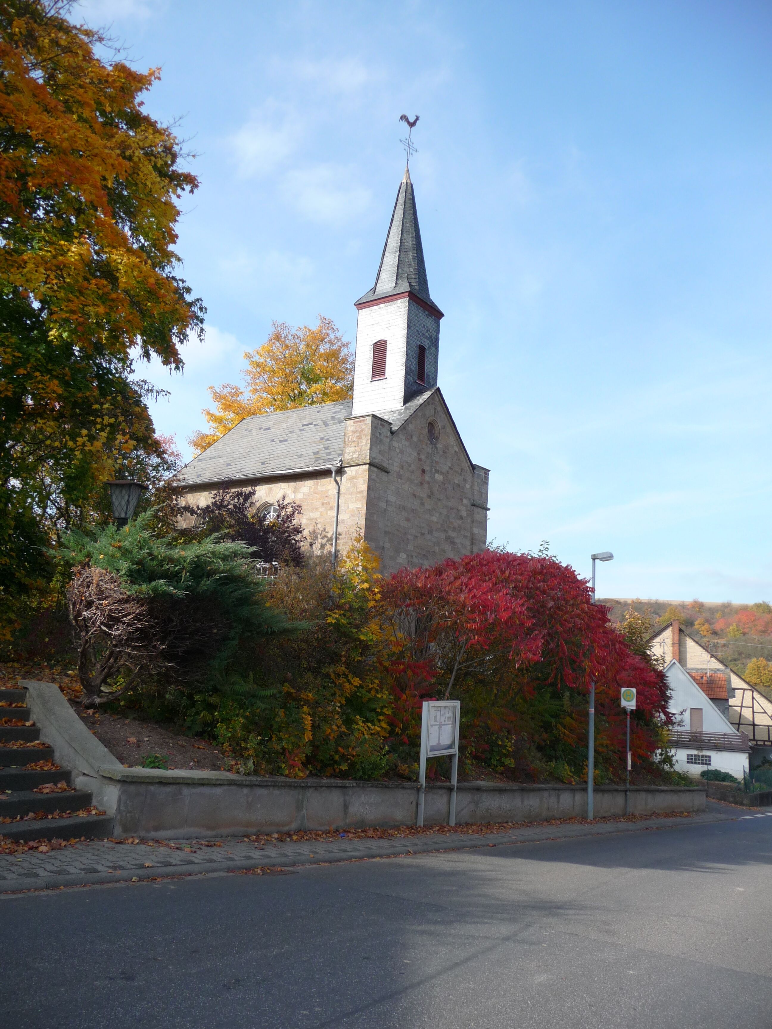 Uma igreja com um campanário e um indicador de vento, cercada por folhagem outonal e uma estrada pavimentada.